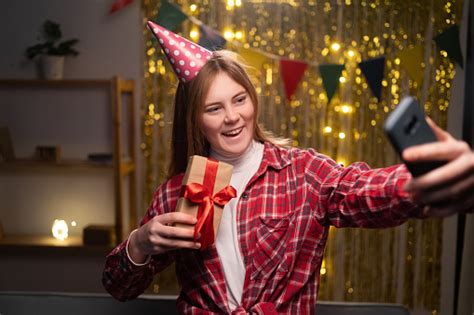 먼 의사 소통과 축하 휴대 전화를 들고 웹캠에 선물을 보여주고 파티 모자를 쓰고 집에서 가상 생일을 축하하는 백인 소녀의 초상화 원격 생일 18 19세에 대한 스톡 사진 및