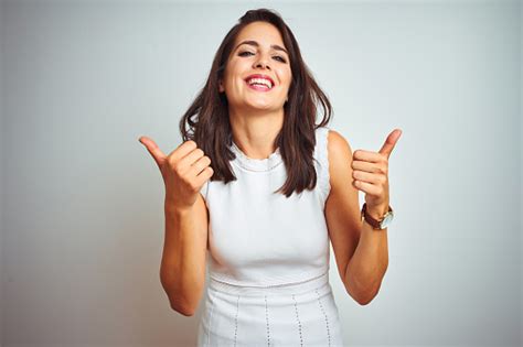 흰색 고립 된 배경 성공 표지판 위에 서있는 드레스를 입은 젊은 아름다운 여성은 손으로 긍정적 인 제스처를하고 엄지 손가락으로