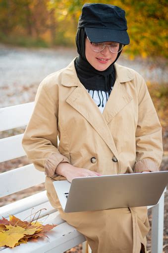 베이지색 트렌치코트를 입은 무슬림 여성이 공원에 커피 한 잔과 노트북을 들고 벤치에 앉아 있다 무슬림 비즈니스 우먼 학생 시력을 위한 안경 20 24세에 대한 스톡 사진 및