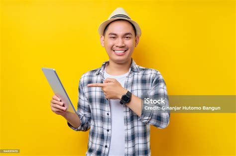 캐주얼 옷 모자를 쓴 웃는 여행자 관광객 아시아 남자가 노란색 배경에 고립된 태블릿 Pc 컴퓨터를 손가락으로 가리키고 있다