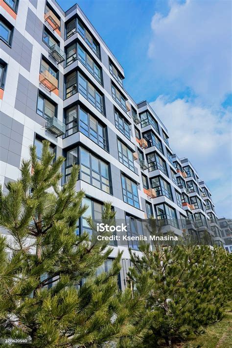 회색과 흰색 톤의 다층 건물로 현대 건축의 주황색 인서트가 있으며 대형 창문과 에어컨 바구니가 있으며 구름이있는 푸른 하늘을 배경으로 침엽수 사이에 서서 아래에서 볼 수