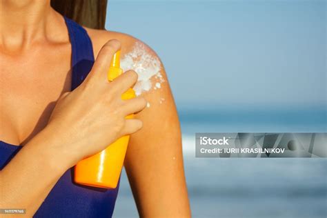 파란색 수영복을 입은 젊은 여성은 플라스틱 오렌지색 패키지 스프레이 병을 들고 몸에 자외선 차단제를 바르고 있습니다 해변에서