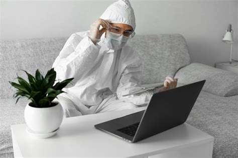 보호 흰색 정장과 의료 마스크를 가진 남자는 코로나 바이러스 전염병으로 인해 노트북과 태블릿소파에서 집에서 일하고 있습니다