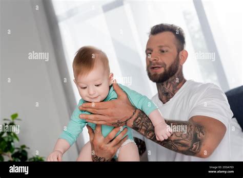 Selective Focus Of Bearded Tattooed Man Holding Baby Boy In Bedroom