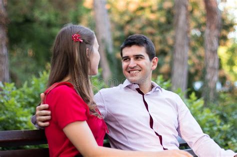 Pareja Joven Enamorada Sentada En Un Banco Al Hombre Que Mira A La Mujer Sonriendo Imagen De