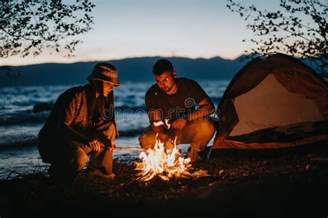 Friends Enjoying A Campfire By The Lake At Sunset Cooking Dinner