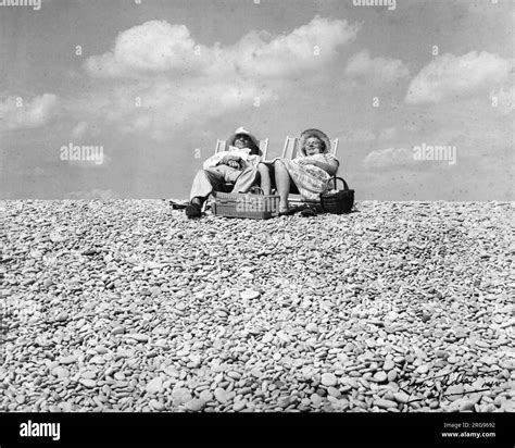 Contented Middle Aged Couple At The Seaside Sitting In Deckchairs On A Pebbly Beach With A
