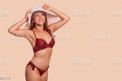 머리에 밀짚 모자를 쓰고 스튜디오 벽에 손을 들고 포즈를 취하고 미소를 짓고 빨간 수영복을 입고 바다 근처에서 레크리에이션을 하고 있는 슬림한 검은 머리 의 여성의 실내
