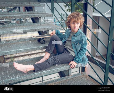 Full Body Of Barefoot Curly Haired Preteen Boy In Jeans Jacket Sitting