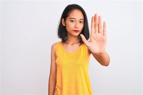 노란 캐주얼 티셔츠를 입은 젊은 중국 여성이 고립된 흰색 배경 위에 서서 손바닥으로 노래를 멈추고 있습니다 얼굴에 부정적이고 심각한 제스처로 경고 표현 T 셔츠에 대한 스톡