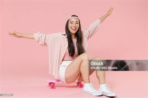 캐주얼 한 옷을 입고 쾌활한 젊은 아시아 여성 소녀의 전체 길이 초상화 파스텔 핑크 배경 스튜디오 초상화에 고립 된 모자 사람들 라이프 스타일 개념 스케이트보드에 앉아 손을