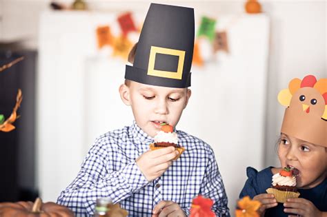 아이들은 축제 테이블에 앉아 추수 감사절을 축하합니다 종이 칠면조 모자와 순례자 모자를 쓴 아이들은 컵케이크를 먹고 우유를 마시는 것입니다 먹기에 대한 스톡 사진 및 기타