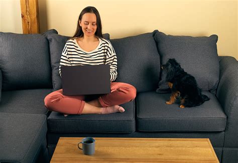 원격 작업에서 여자는 웃는 연꽃 위치에 소파에 앉아 머그잔과 테이블 근처 그녀의 무릎에 노트북에 입력 가까운 에 그녀의 작은 개