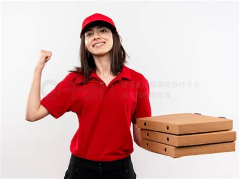 积极高清图片 身穿红色制服、头戴帽子的年轻送货女孩拿着一堆披萨盒，握紧拳头，快乐而积极地微笑着，站在白色背景上 餐饮美食免费下载格式