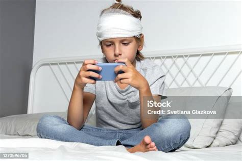 머리 부상과 붕대를 가진 작은 백인 금발 소년은 침대에 앉아 휴대 전화를 사용하고 있습니다 사고 후 복구 원격 통신 기술 및