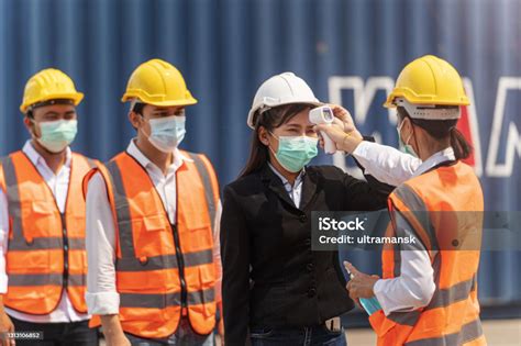 얼굴 의료 마스크와 안전 드레스를 입은 공장 여성 근로자는 비접촉 적외선 온도계로 대기열에 서있는 근로자의 온도를 측정합니다