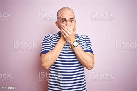 분홍색 고립 된 배경 위에 캐주얼 스트라이프 블루 티셔츠를 입고 수염을 가진 젊은 대머리 남자는 실수로 손으로 입을 덮는 충격을