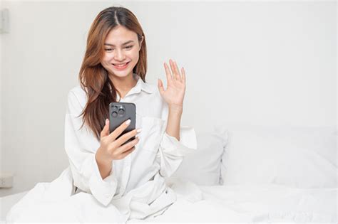 하얀 잠옷을 입은 여자가 주말 아침에 일어나 휴식을 취하고 노트북 휴대폰을 가지고 놀며 하얀 침실 안의 유리에서 빵을 먹고 차를