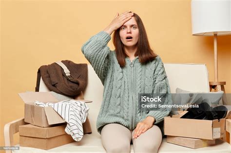 베이지색 벽에 옷을 입힌 상자 사이에 소파에 앉아 큰 눈과 입을 크게 벌린 채 카메라를 바라보는 손바닥 제스처를 보여주는 충격을
