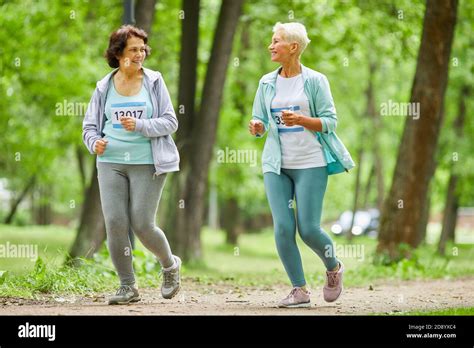 Full Body Shot Of Two Active Senior Women Running Marathon Race In City Park Talking To Each