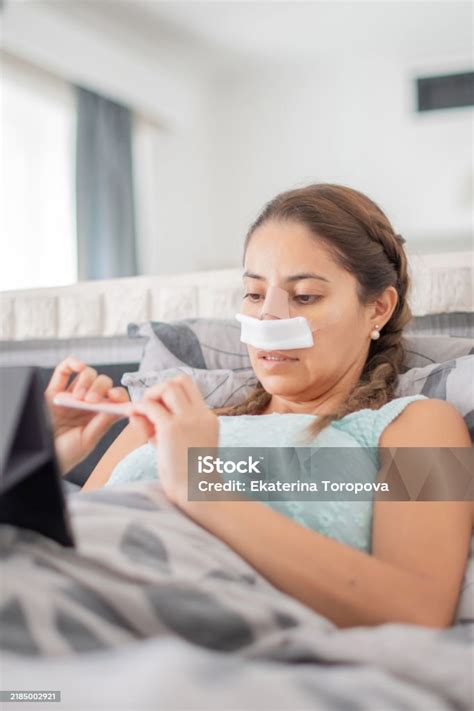 여자는 코 성형 수술 후 캐주얼 한 옷차림으로 얼굴에 붕대를 감고 침대에 누워 손톱을 정리하고 태블릿으로 좋아하는 영화를 봅니다 개성 개념에 대한 스톡 사진 및 기타 이미지