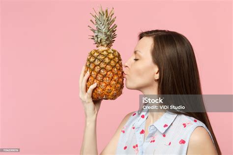 여름 옷에 젊은 여자의 측면 보기 스튜디오에서 분홍색 파스텔 벽 배경에 고립 된 신선한 잘 익은 파인애플 과일 키스를 개최 합니다 사람들은 생생한 라이프 스타일 휴식 휴가