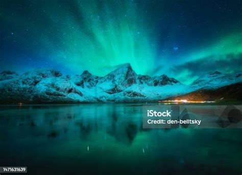 눈 덮인 산 위의 북극광 얼어붙은 바다 노르웨이 로포텐의 밤에 물에 반사 오로라 보리 얼리 스와 눈 덮인 바위 극지방의 불빛 별이 빛나는 하늘 피요르드가 있는 겨울 풍경