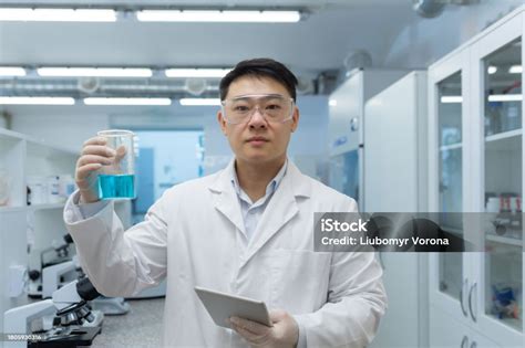 젊은 아시아 남성 과학자 화학자가 손에 플라스크와 파란색 액체 태블릿을 들고 실험실에 서 있는 초상화 그는 진지한 표정으로 카메라를 가리킨다 35 39세에 대한 스톡 사진
