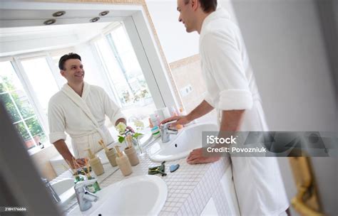 하얀 목욕 가운을 입은 남자가 욕실 거울에 비친 자신을 보며 미소를 짓고 있다 깨끗하고 현대적인 욕실에는 타일로 마감된 조리대 식물 세면도구 밝고 햇살이 잘 드는 분위기가