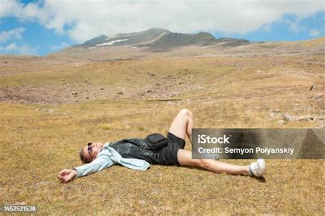 데님 재킷과 반바지를 입은 청년이 산의 잔디밭에 누워 팔을 옆으로 벌렸다 휴가 중에 탈출 한 남자가 산에서 자연 속에서 인생을 즐기게되어 기쁩니다 사람들에 대한 스톡 사진
