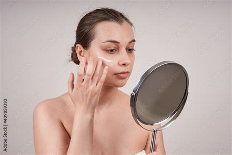 Cute Young Topless Woman Applying Nourishing Cream To Her Face While Looking In The Mirror
