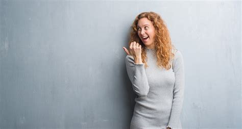 고 행복 얼굴 미소와 측면까지 손가락으로 보여주는 회색 그런 지 벽에 빨강 머리 젊은 여자 가리키기에 대한 스톡 사진 및 기타