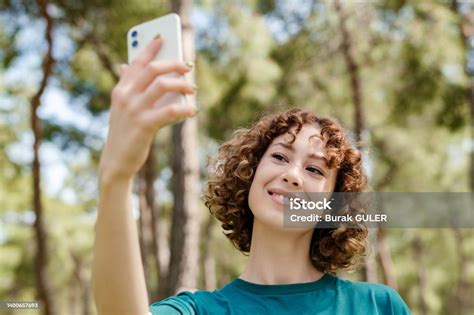 녹색 캐주얼 한 옷을 입고 도시 공원에 서있는 쾌활한 여성 스마트 폰으로 자화상을 찍는 야외 활동 그녀는 화면을보고 셀카를