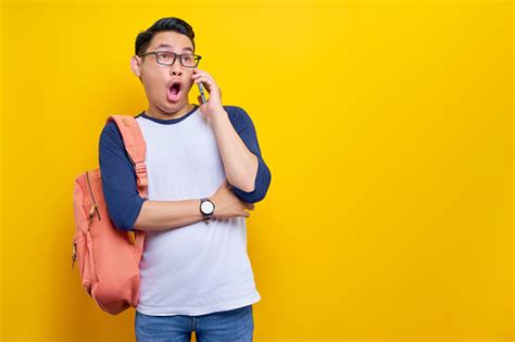충격을 받은 젊은 아시아 남자 학생은 캐주얼 옷 배낭 안경을 쓰고 친구와 휴대전화로 이야기를 나누며 노란색 배경에 고립된 채