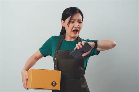 앞치마를 두른 아시아 여성은 시간을 확인하고 판지 상자를 들고 충격을 받고 배경 위에 고립되어 서 있다 소기업에 대한 스톡 사진