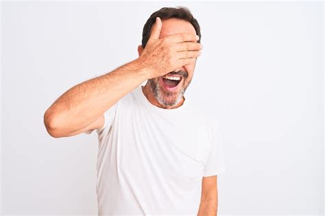 고립된 흰색 배경 위에 서있는 캐주얼 한 티셔츠를 입은 중년 잘생긴 남자는 놀라움을 위해 눈을 가리는 얼굴에 손으로 웃고