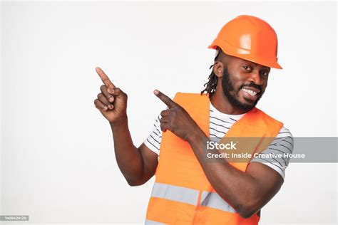 반사 오렌지 조끼와 하드 햇을 입은 젊은 아프리카 계 미국인 남자 건설 노동자 엔지니어 건축가는 흰색 배경에 고립 된 여유 공간을 보여주는 복사 공간을 가리 킵니다