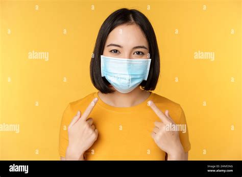 Portrait Of Smiling Asian Woman In Medical Face Mask Pointing At Her