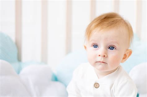 6개월 된 아기의 클로즈업 초상화 흰색 면 바디수트를 입은 밝은 침실의 유아용 침대에서 파란 눈을 가진 금발 소년 아동용품의