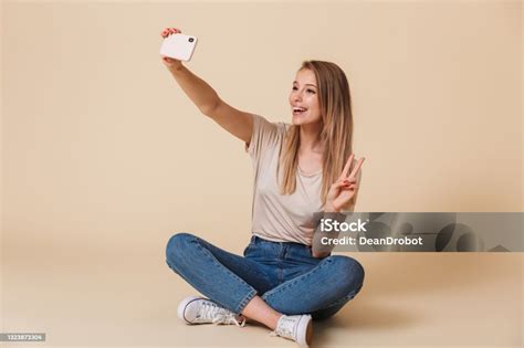 평화 표지판을 보여주는 캐주얼 한 옷을 입고 다리가 교차 바닥에 앉아있는 동안 휴대 전화로 셀카 사진을 찍는 행복한 젊은 여성이 베이지 색 벽을 통해 고립되었습니다 20