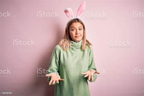 고립 된 분홍색 배경 에 서 부활절 토끼 귀를 입고 젊은 아름다운 여성은 팔과 손을 들고 단서와 혼란 스러운 표정 의심 개념