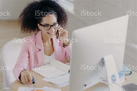 행복 곱슬 머리 여성의 사진은 광학 안경을 착용 분홍색 재킷 컴퓨터 디스플레이에 주의 깊게 보이는 열린 메모장바탕에 앉아 스마트