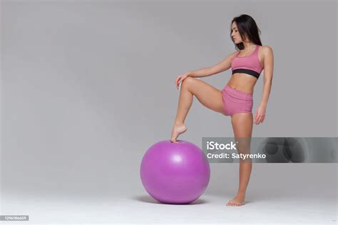 현대 핑크 스포츠웨어를 입은 젊은 매력적인 소녀가 회색 배경에 고립된 피트니스 볼에 발을 디디는 포즈를 취하고 있습니다 전체 길이 가냘픈에 대한 스톡 사진 및 기타 이미지
