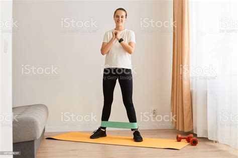 흰색 티셔츠와 검은 색 레긴을 입고 운동 서서 다리를 위한 운동을하고 훈련을 위해 저항 밴드를 사용하여 운동하는 스포티 한