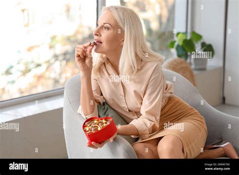 Mature Woman Eating Heart Shaped Chocolate Candies In Armchair At Home