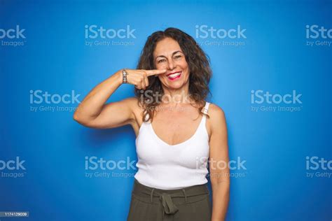 중년 시니어 여성은 파란색 고립 된 배경 위에 서있는 곱슬 머리를 가진 손 손가락을 얼굴과 코로 가리키며 쾌활한 미소를 짓고 있습니다 뷰티 컨셉 가리키기에 대한 스톡 사진