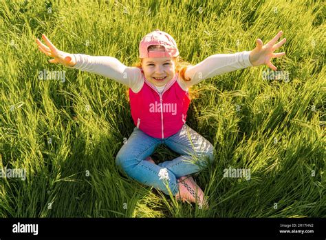 A Cheerful Girl Sits In The Thick Green Grass With Her Hands Up In The
