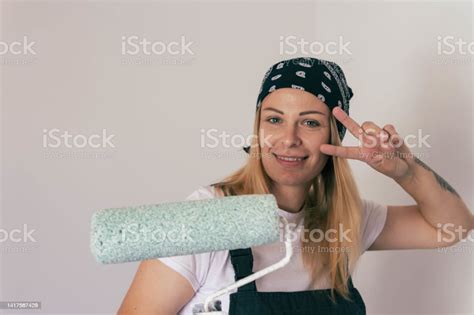 반다나에 금발 머리를 가진 유럽 외모의 소녀는 집에서 롤러로 벽을 흰색으로 칠합니다 카메라를 쾌활하게 마주합니다 비문을위한 장소가 있습니다 고품질 사진 손에 대한 스톡 사진