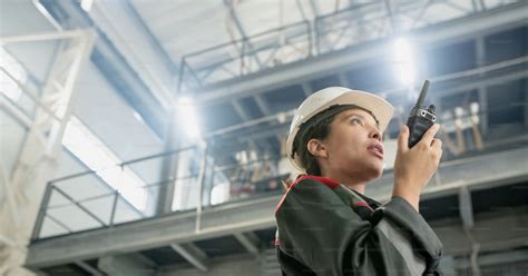 현대 공장의 대규모 작업장에 서 있는 동안 워키토키를 사용하는 보호 헬멧과 작업복을 입은 젊은 여성 엔지니어 사진