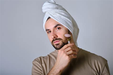 얼굴에 괄사 롤러를 사용하여 머리에 수건을 쓰고 카메라를 바라보는 잘생긴 청년의 중간 장면 수건에 싸다에 대한 스톡 사진 및